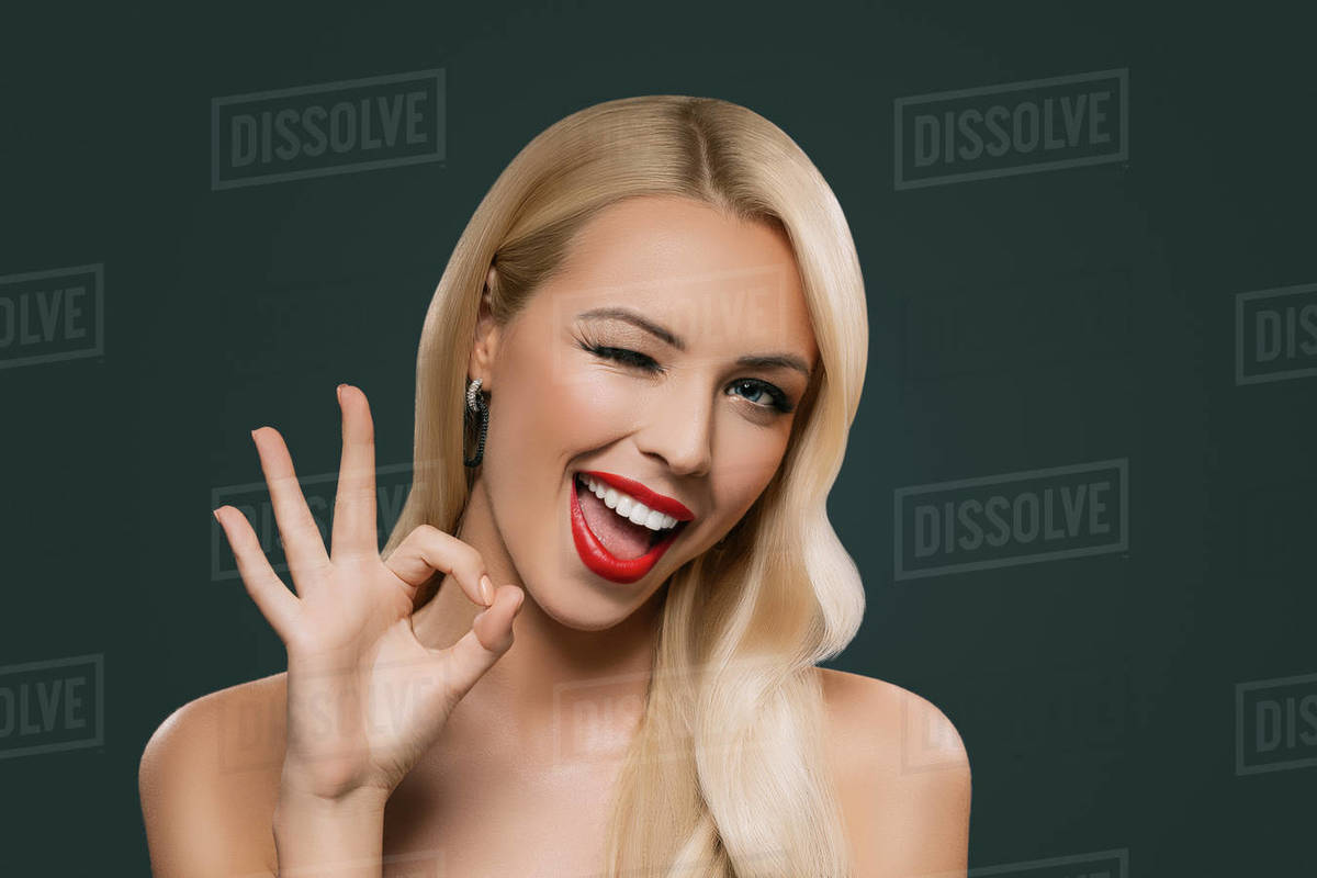 Attractive blonde winking woman showing okay sign, isolated on grey ...