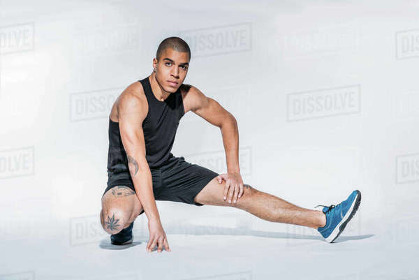 african american sportsman stretching legs and looking at camera ...