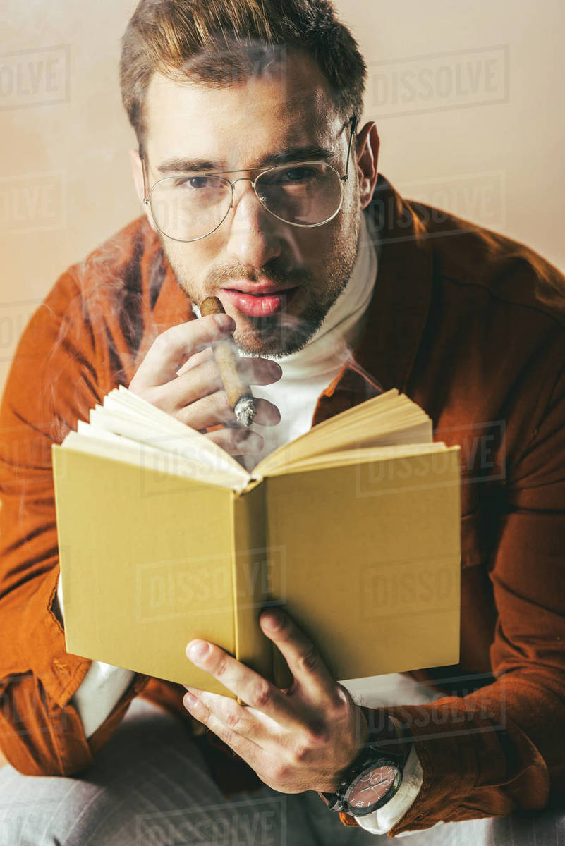 Portrait of young man with cigar and book in hands looking at camera ...