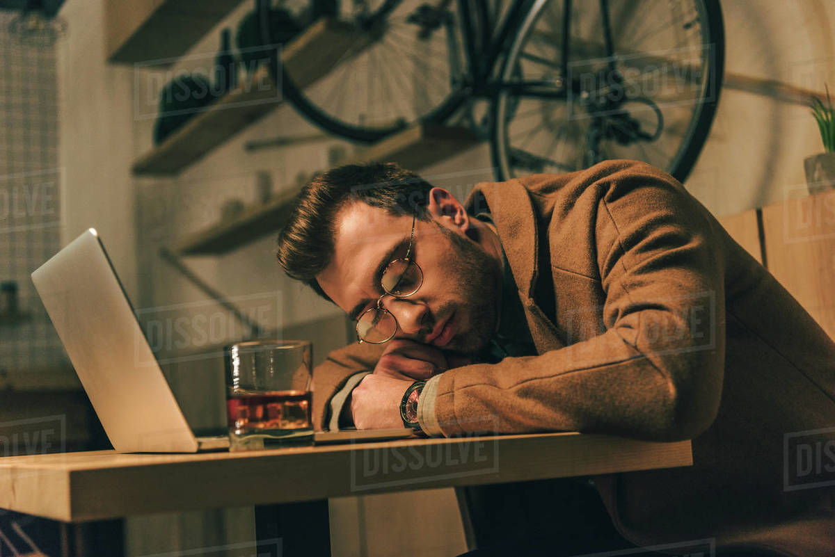 Tired man sleeping on table with glass of whiskey and laptop in cafe ...