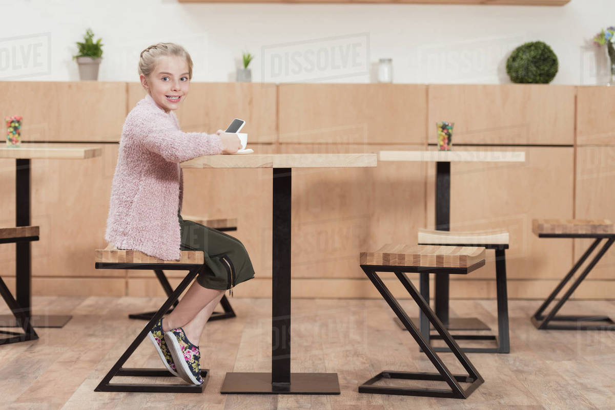 smiling child sitting at table with smartphone and looking at camera ...