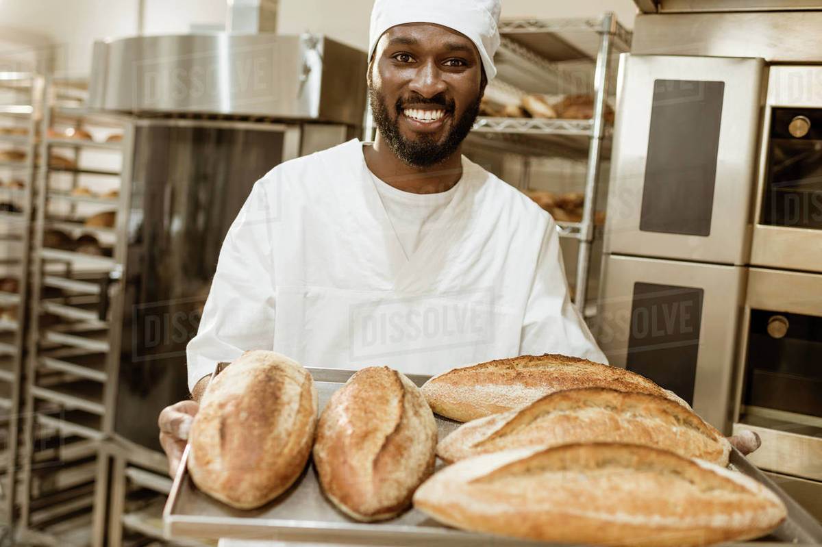 Handsome african american baker with tray of fresh loaves of bread on ...
