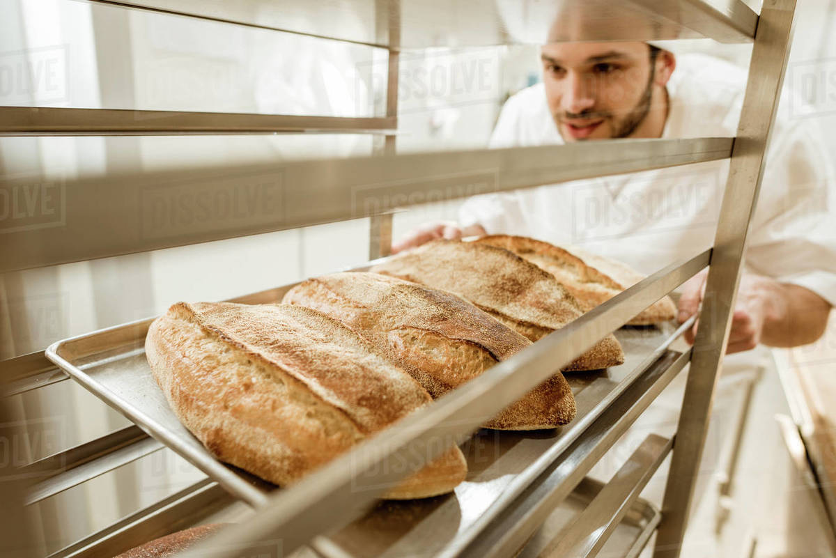 Close-up shot of handsome baker putting trays of fresh bread on stand ...