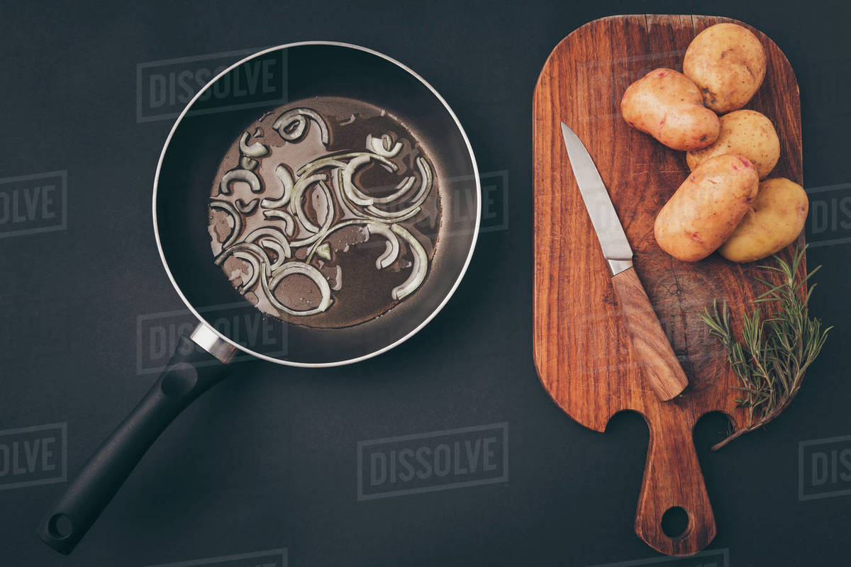 Top view of frying pan with onion and wooden board with potatoes on ...