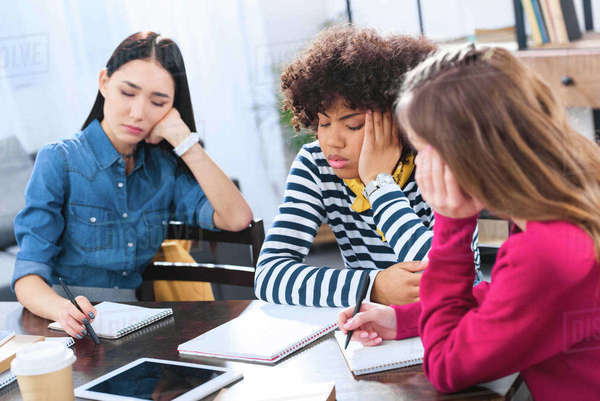 Bored multiracial students sitting at table while doing homework ...