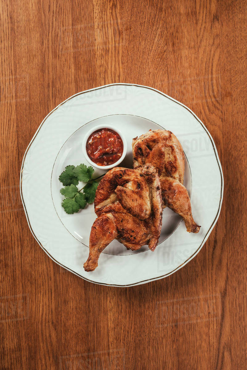 Grilled chicken laying on plate with red sauce in saucer over wooden ...