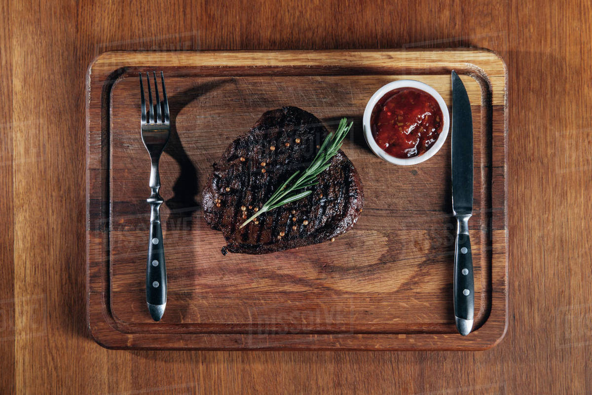 Top view of grilled steak with cutlery and sauce on wooden board ...