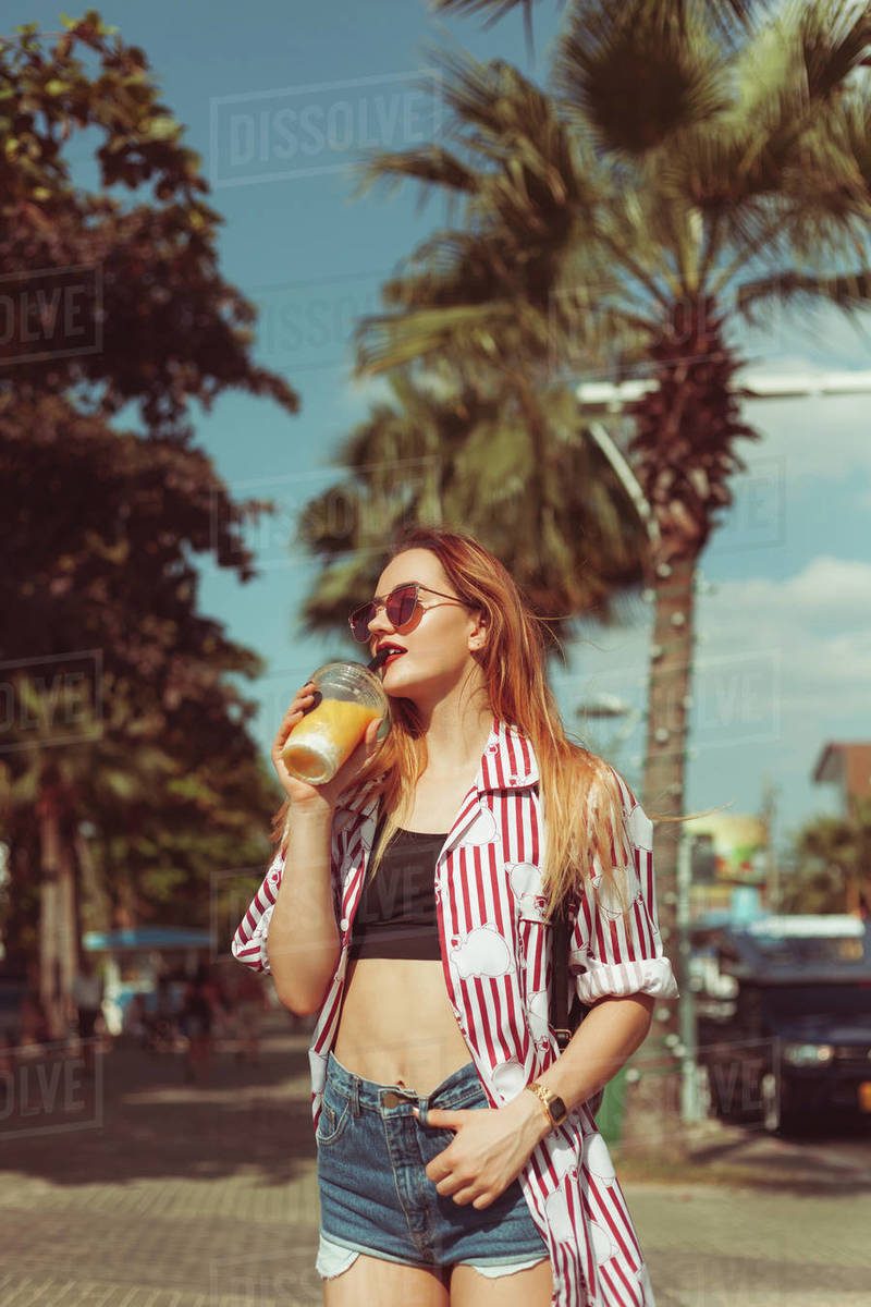Smiling young woman drinking mango shake on sunny street with palm ...