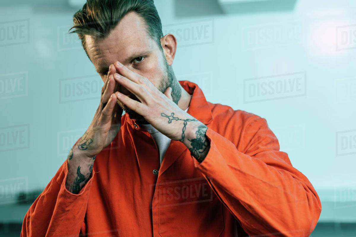 prisoner with tattoos covering face with hands and looking at camera ...