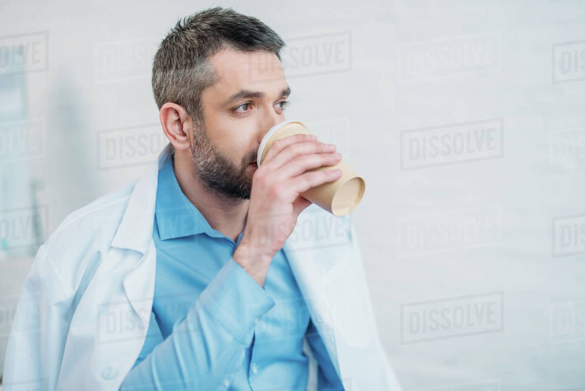 Exhausted doctor drinking coffee from paper cup and looking away ...