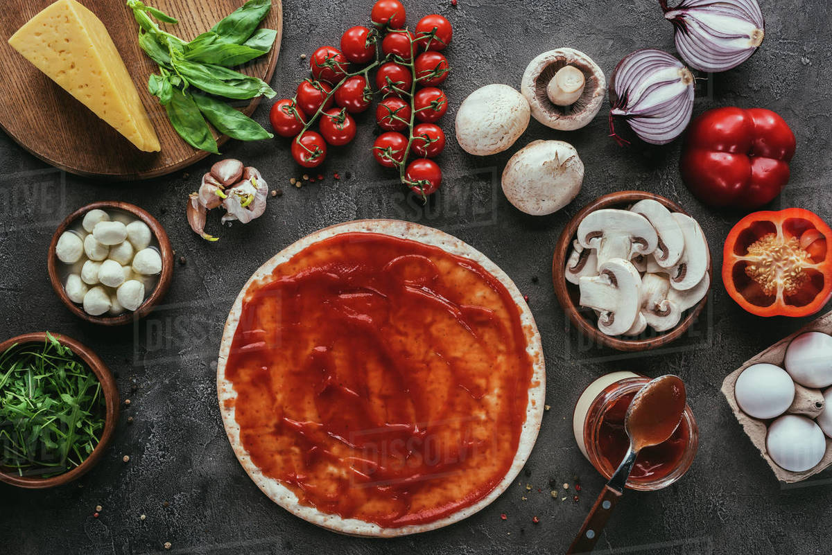 Top view of uncooked pizza dough with sauce and vegetables on concrete ...