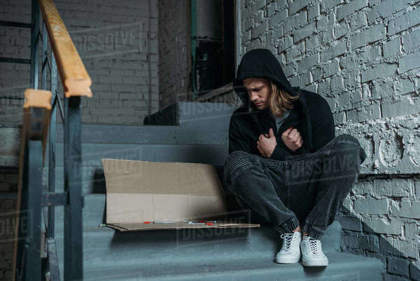 Hooded heroin addicted junkie sitting on stairs with syringes on ...