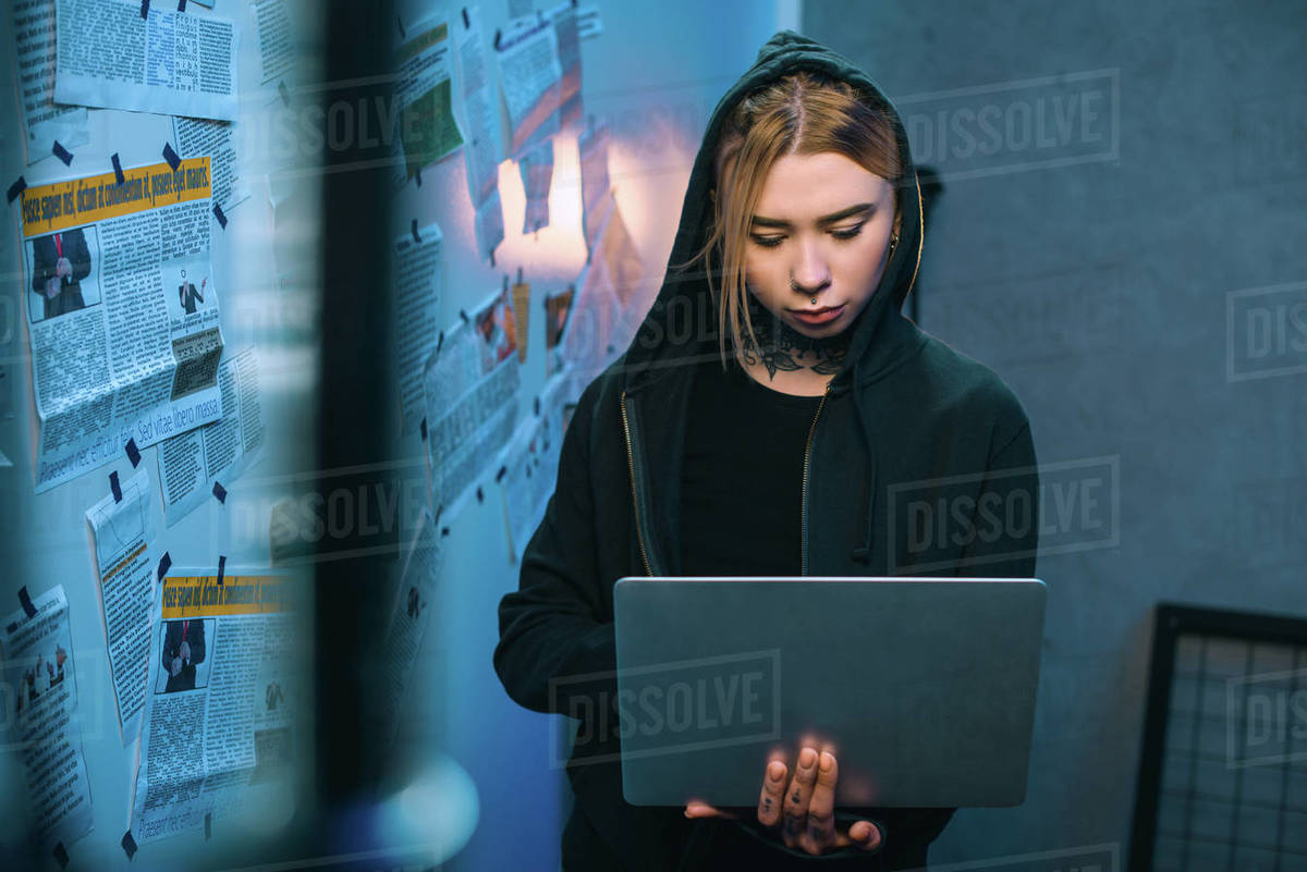 Young female hacker with laptop standing in front of wall with ...