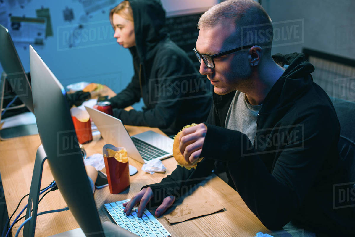 Male hacker working on malware with accomplice and eating junk food ...