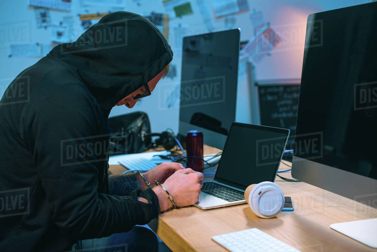 Handcuffed hacker in hoodie in front of laptop at workplace - Royalty ...