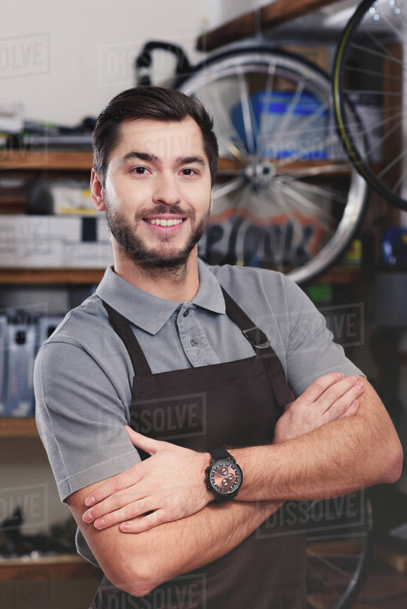 Handsome young bicycle mechanic in apron standing with crossed arms and ...