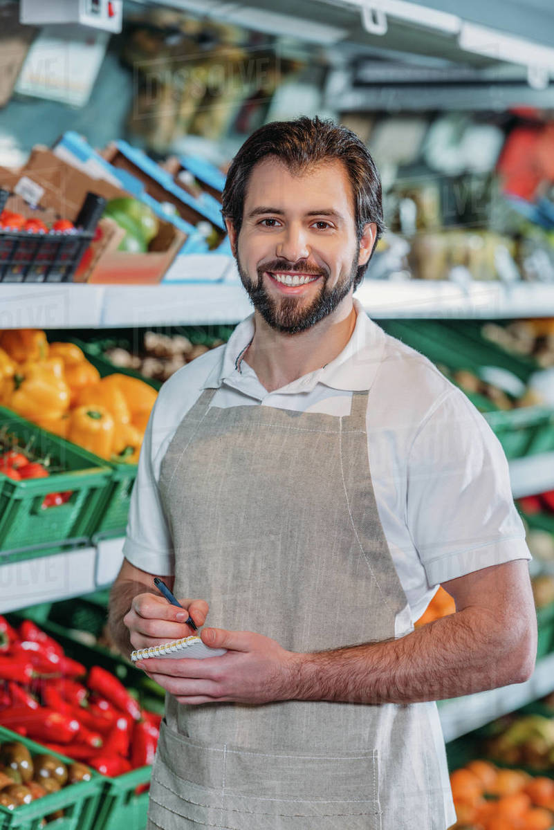 Portrait of smiling shop assistant in apron with notebook in ...