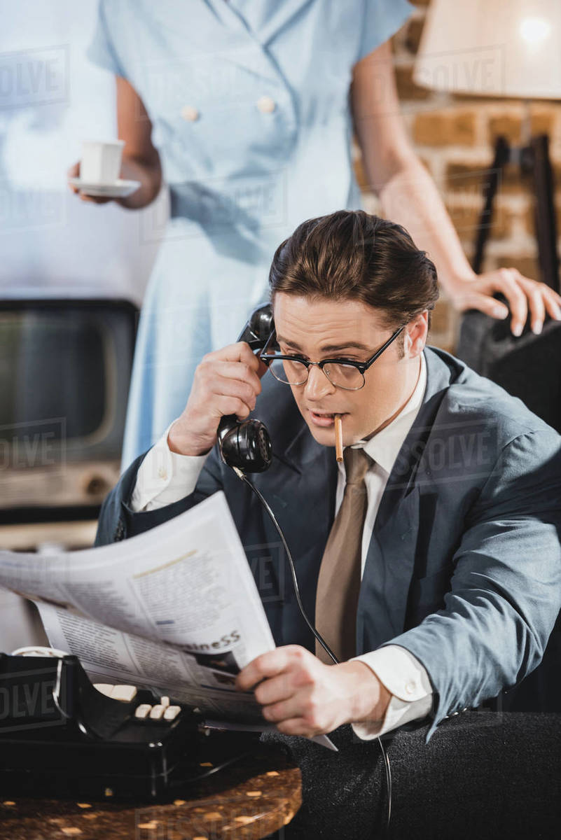 Man with cigarette reading newspaper and talking by vintage telephone ...