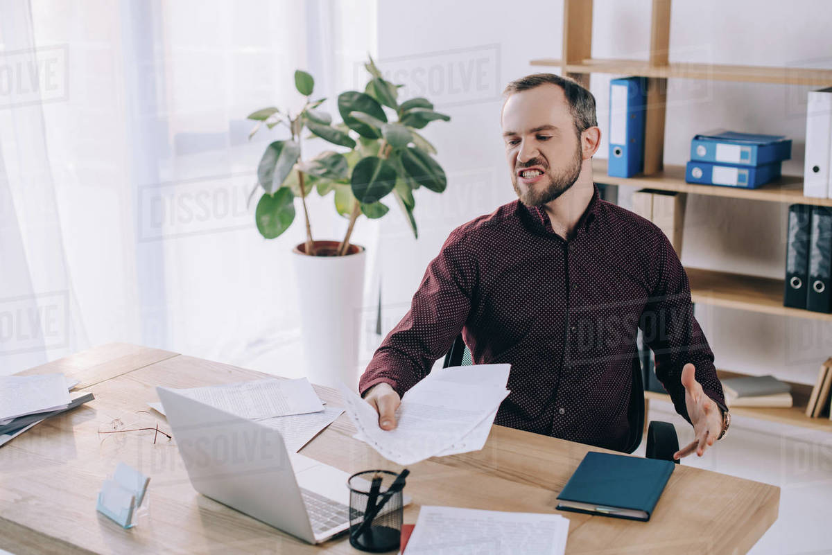 Angry and overworked businessman with papers at workplace with laptop ...