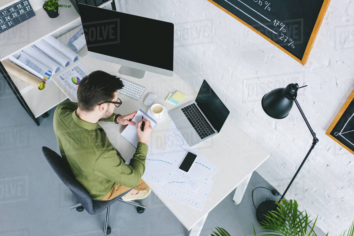 Young man looking at computer screen and taking notes in light office ...