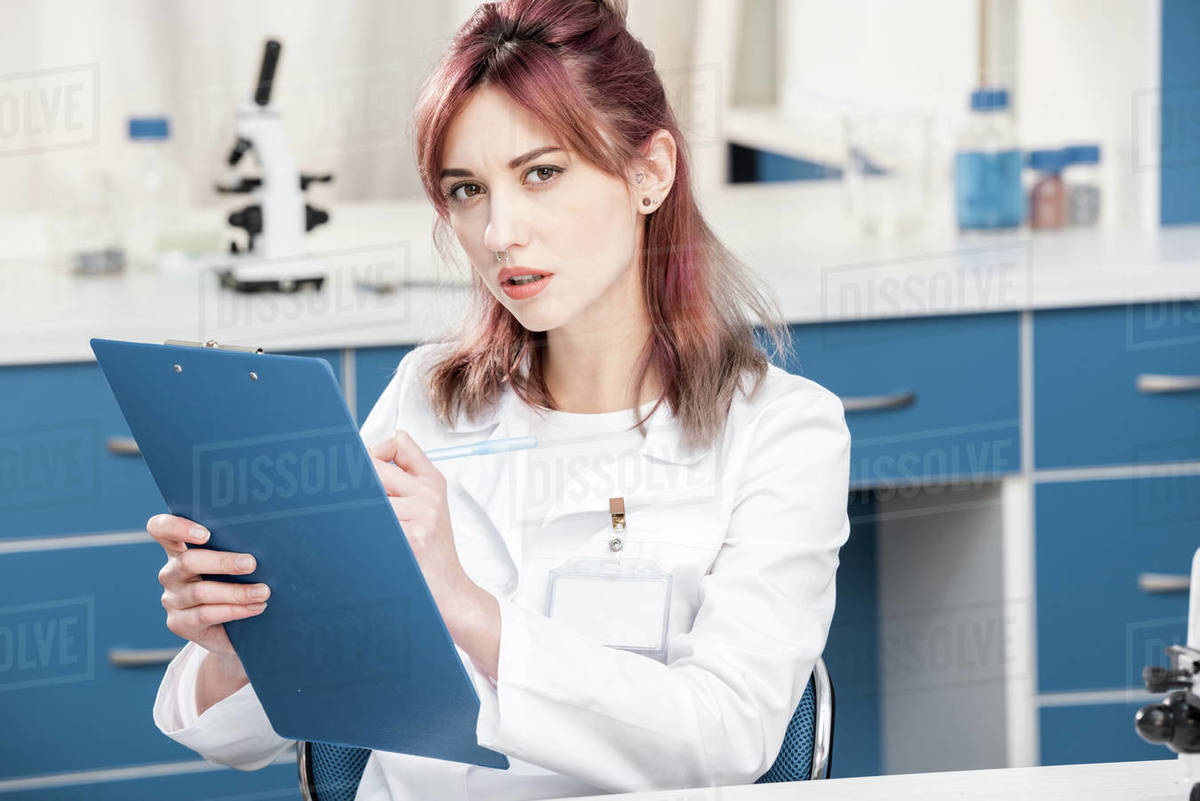 young scientist in lab coat writing in folder and looking at camera in ...