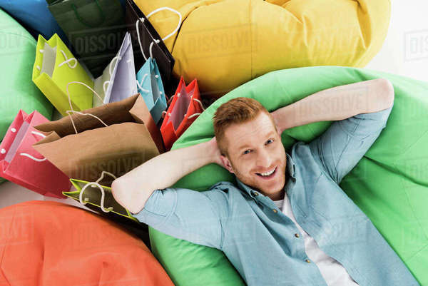 handsome smiling man resting in bean bag chair with colorful shopping ...
