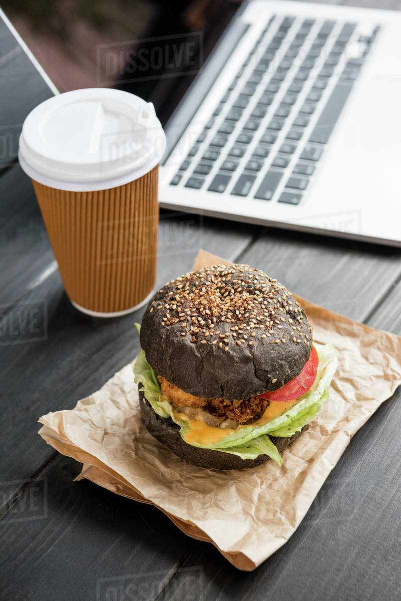 Close up of burger with coffee to go and laptop on wooden tabletop ...