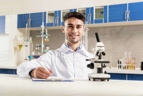 Young smiling lab technician in white coat sitting at the table in ...
