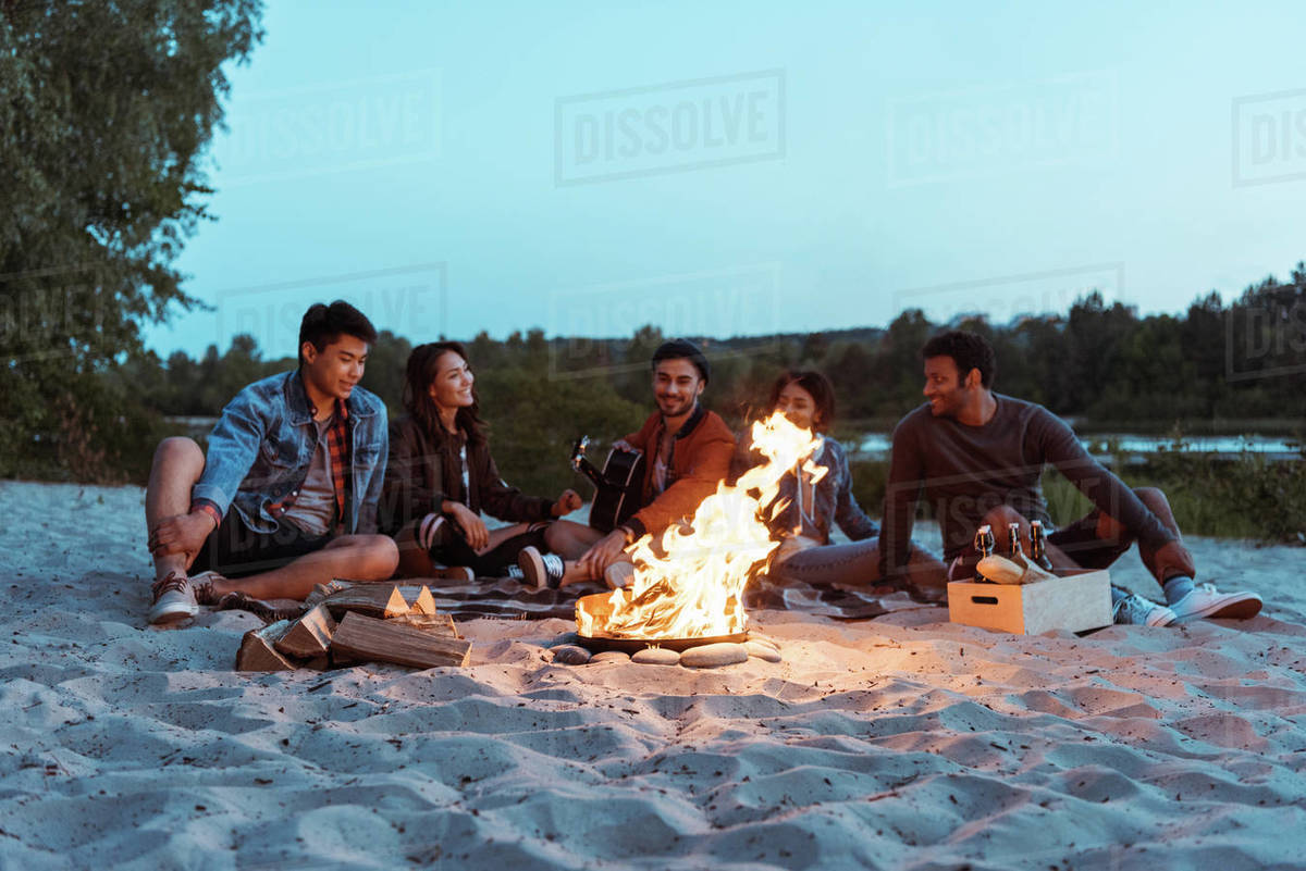 Young multiethnic friends resting near campfire on sandy beach - Royalty-free Stock Photo | Dissolve