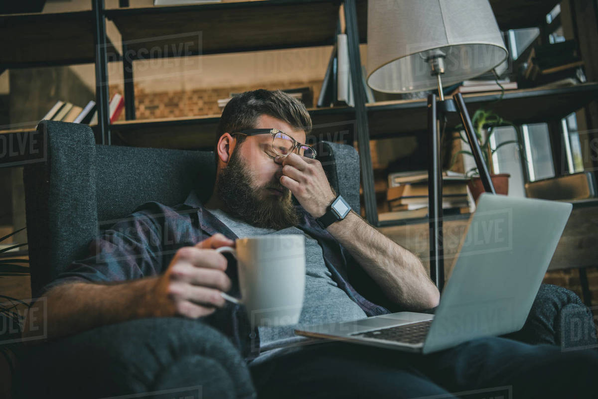 Tired bearded freelancer in eyeglasses using laptop and drinking coffee ...