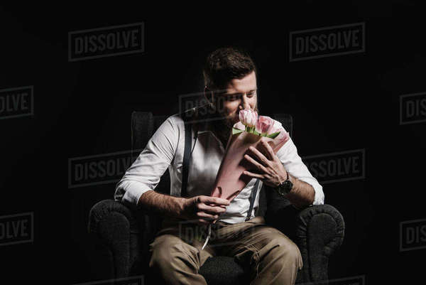 Fashionable man sniffing tulip bouquet while sitting in armchair ...