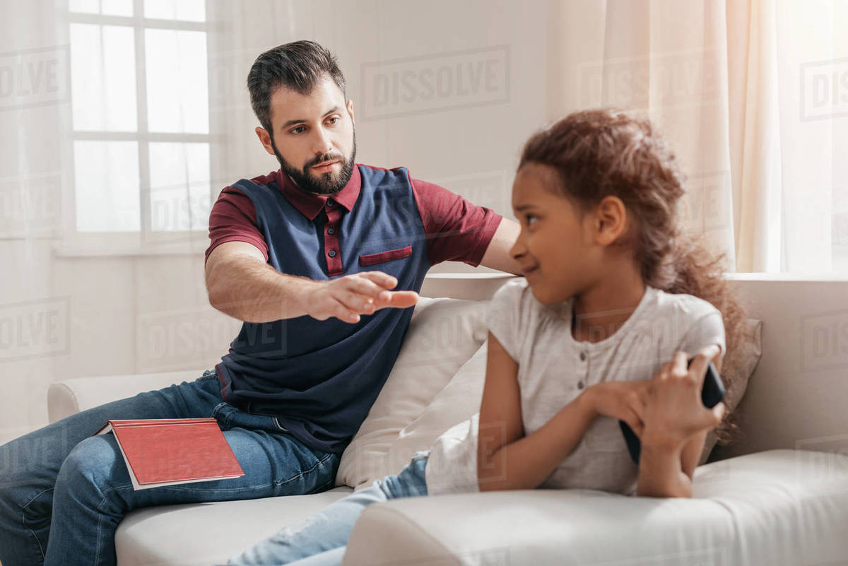 Serious father looking at little daughter holding smartphone at home ...