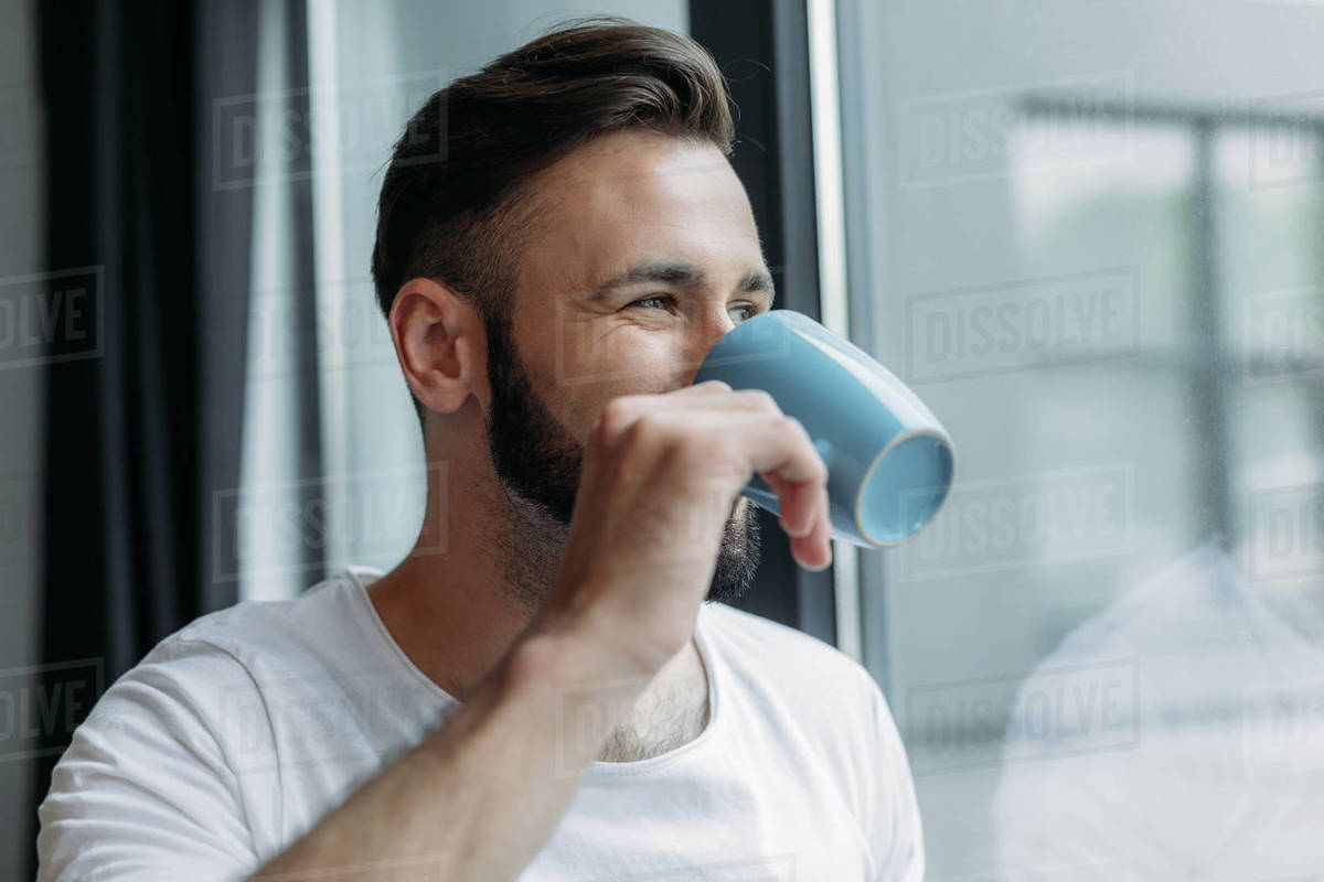 Handsome young man drinking tea from cup and looking at window at home ...