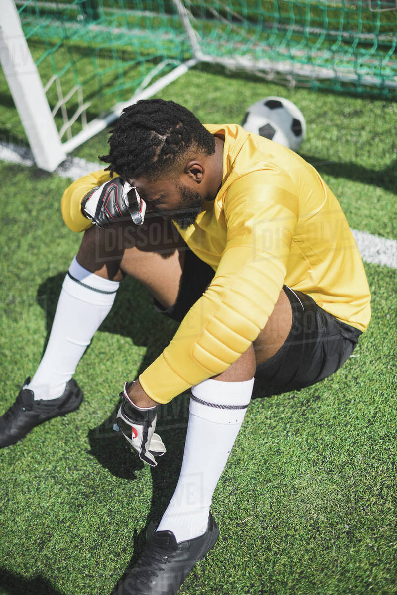Upset african american goalkeeper sitting at goalpost on pitch ...