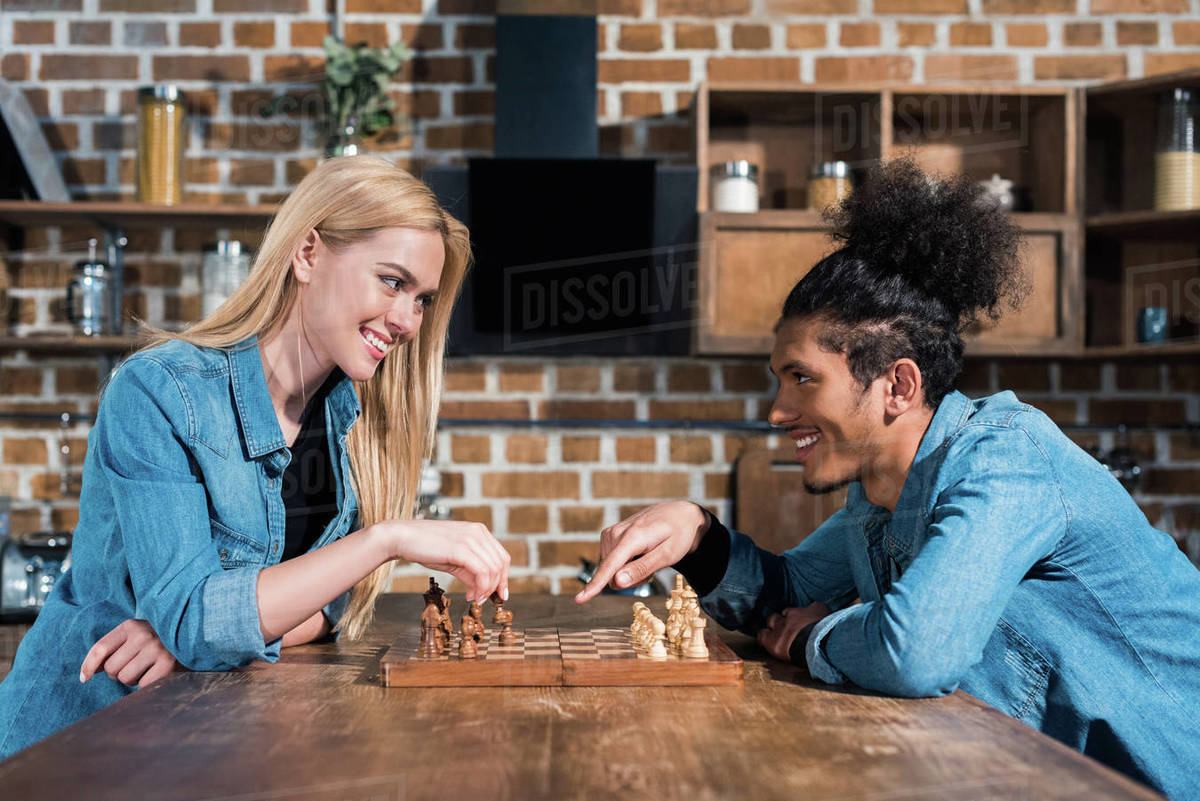 Side view of smiling multiethnic young couple playing chess together in ...