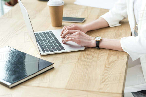 Partial view of businesswoman typing on laptop at workplace with coffee ...