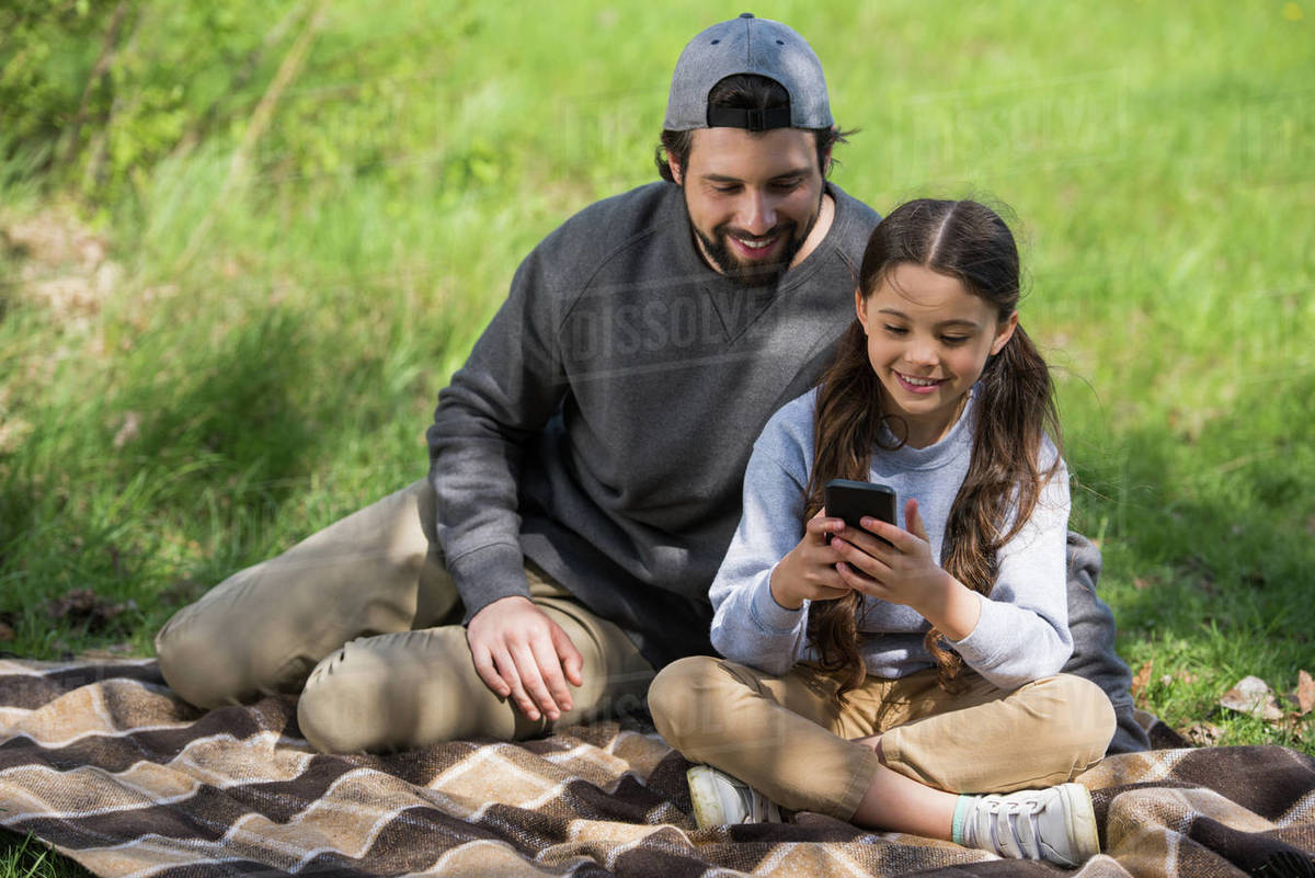Smiling father watching daughter using smartphone on plaid in park ...