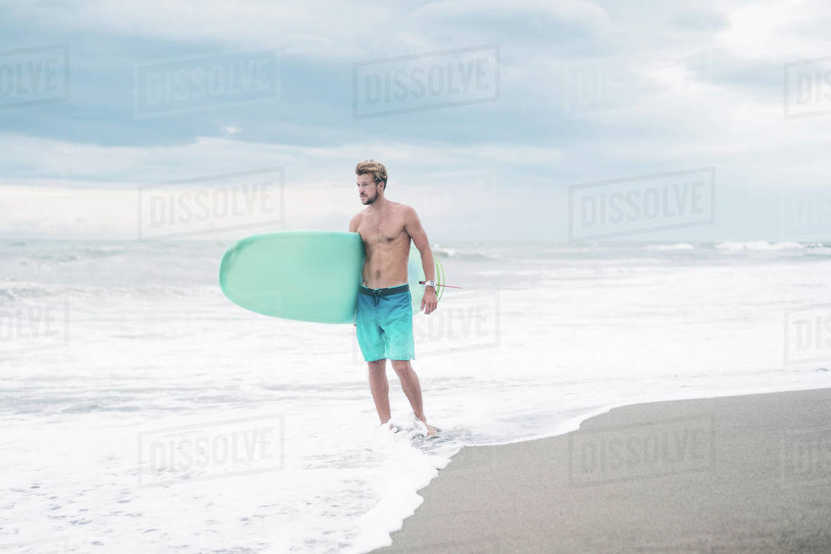 Shirtless surfer standing with surfboard and looking at ocean in Bali ...