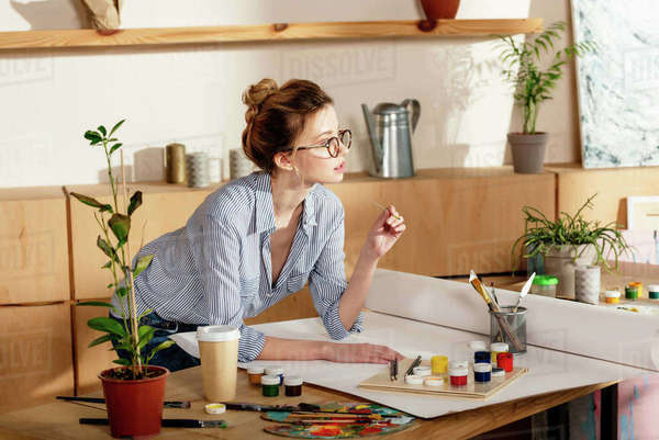 Young female artist in eyeglasses standing at table with empty canvas ...