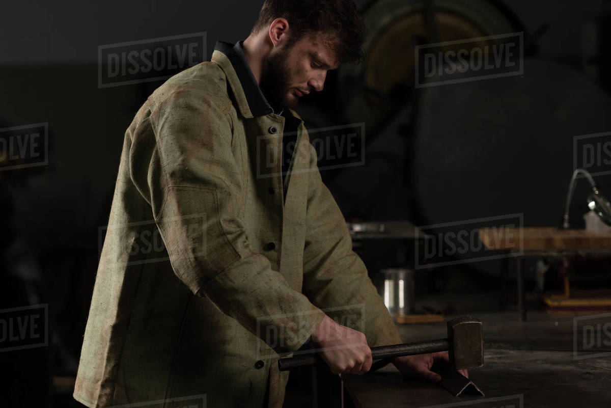 Side view of young workman with hammer and metal part at factory ...