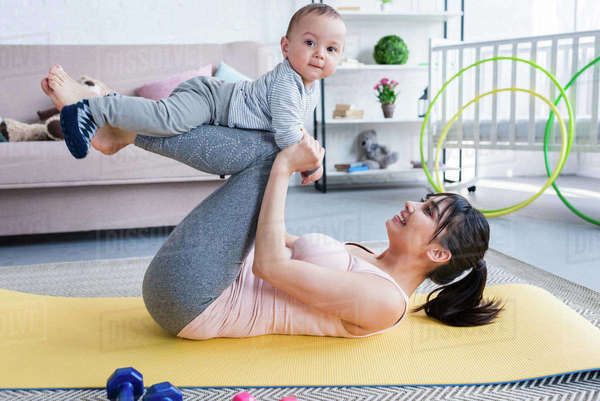 Side view of young beautiful mother working out on yoga mat with her ...