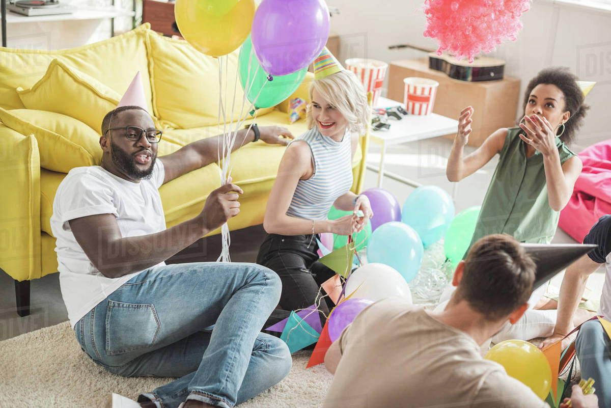 Multicultural friends in party hats sitting on floor with balloons in ...