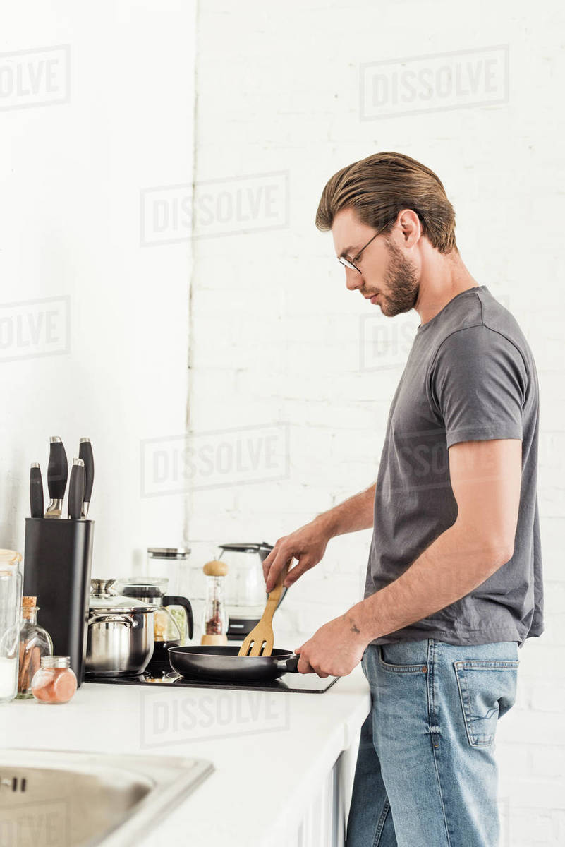 Side view of young man cooking on stove with frying pan and spatula at ...