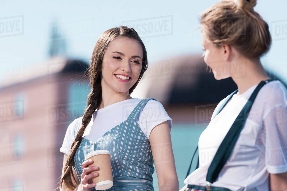 smiling young woman with paper cup of coffee talking to female friend ...