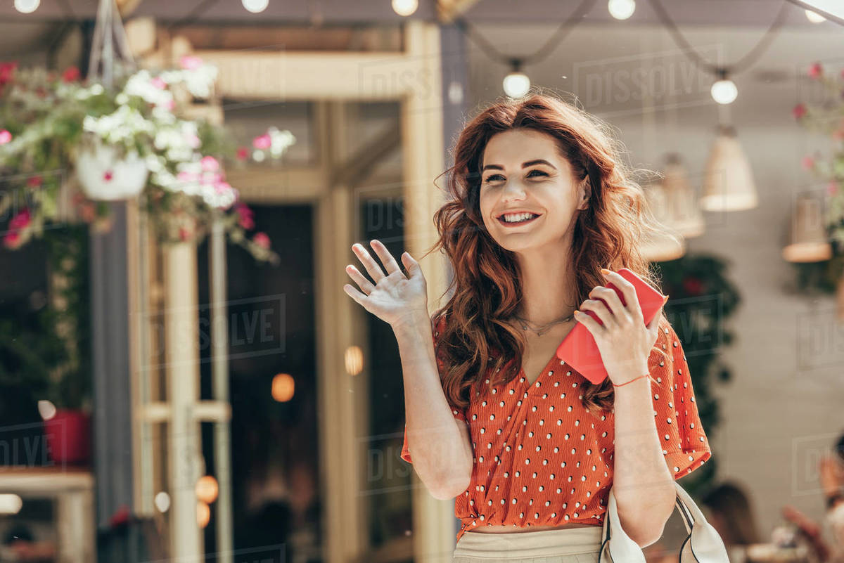 Portrait of cheerful woman with smartphone in hand waiving to someone ...