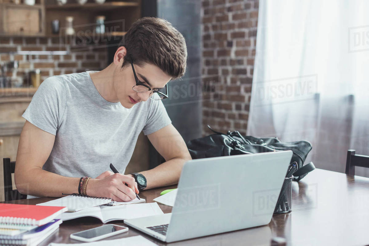 Serious student writing homework at table with laptop and smartphone ...