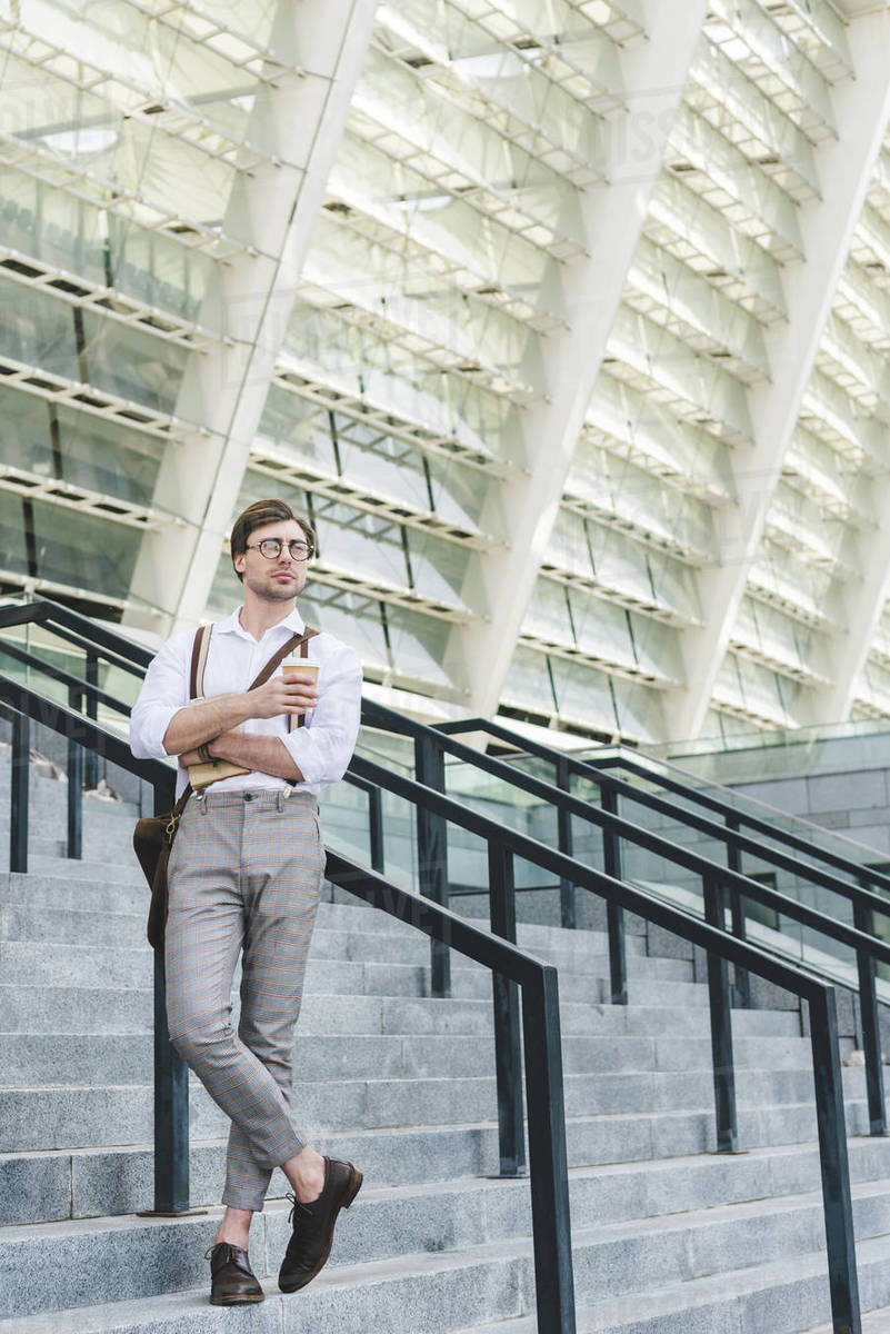 Stylish young man standing on stairs in front of stadium with book and ...