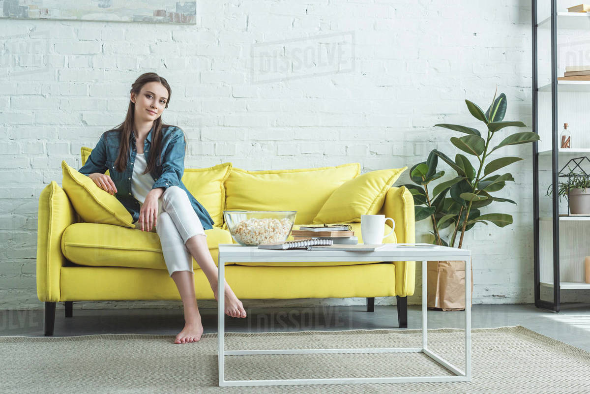 Attractive barefoot girl sitting on sofa and smiling at camera at home ...