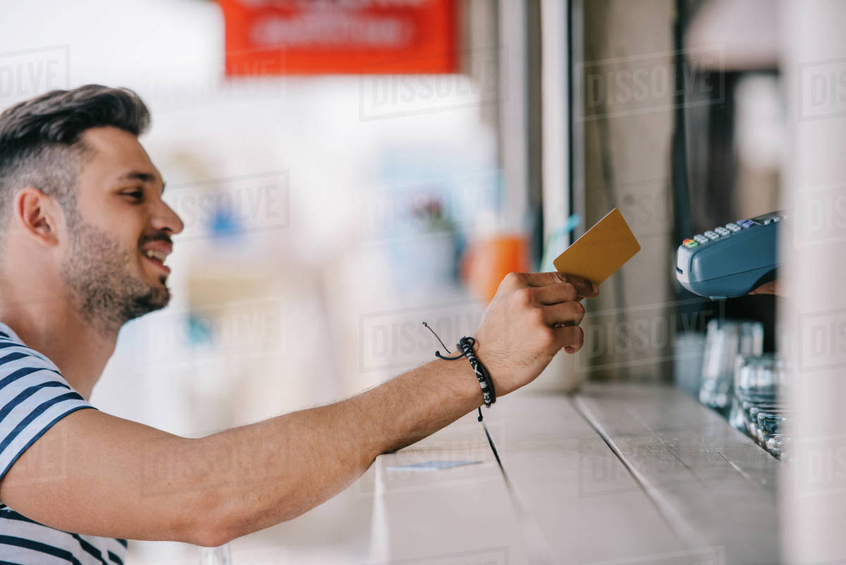 Side view of smiling young man paying with credit card at beach bar ...