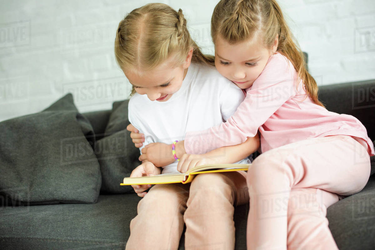 Adorable little sisters reading book together on sofa at home - Royalty ...